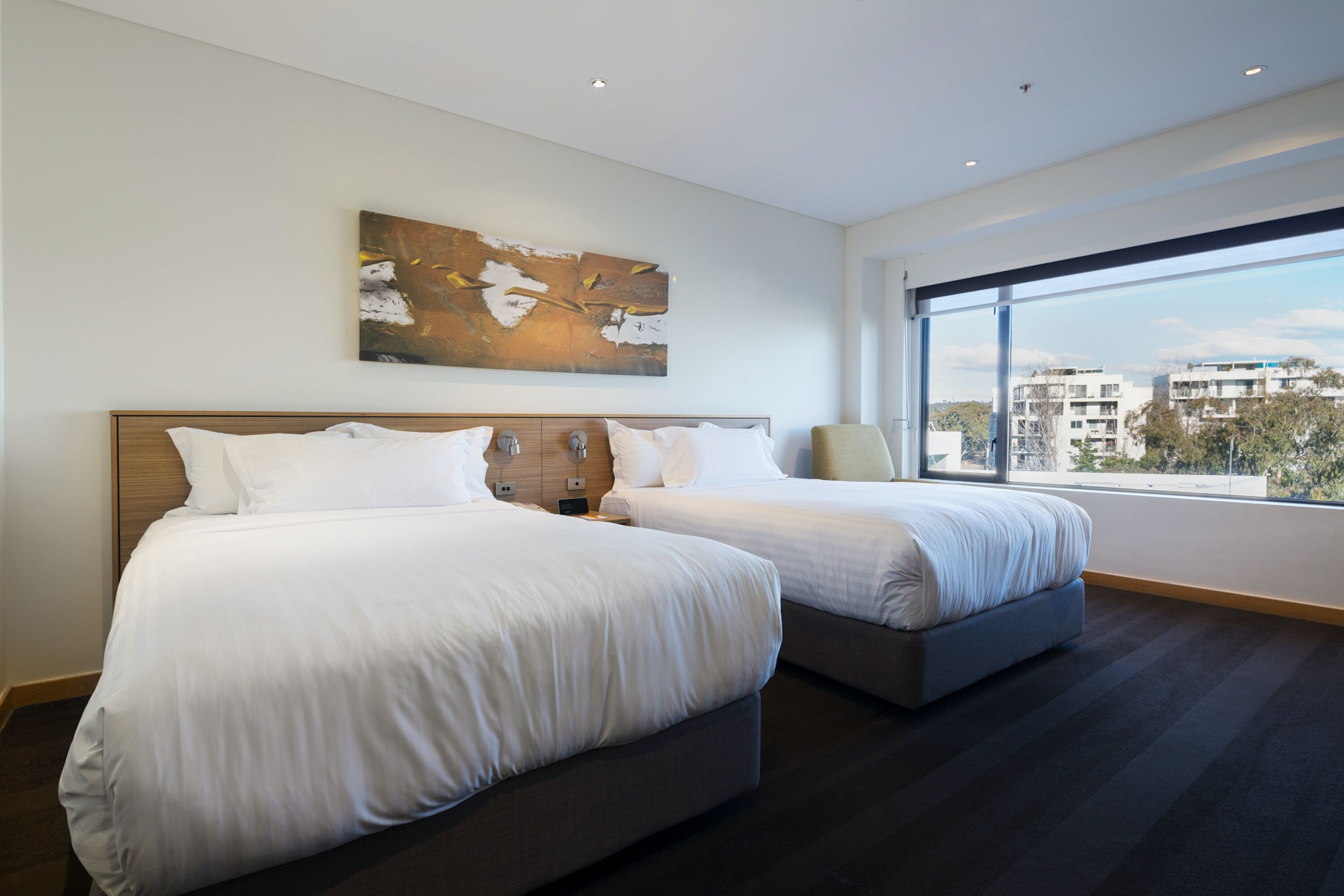 Quarto de hotel com duas camas, uma janela grande com vista para a cidade e uma pintura abstrata moderna na parede.