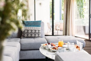 Uma sala de estar moderna com um sofá cinzento, almofadas estampadas, uma mesa de centro redonda com itens de café da manhã e vista para o exterior através de grandes janelas.