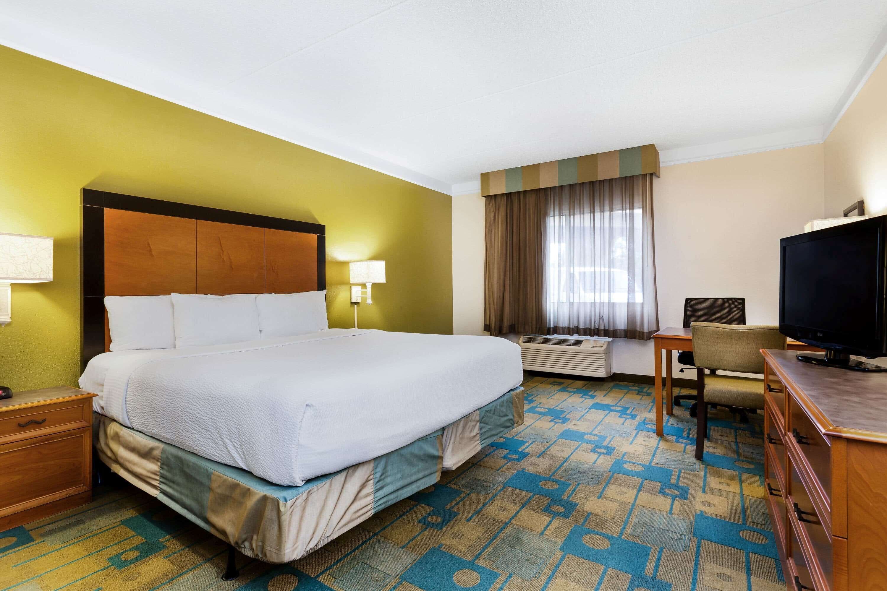 Quarto de hotel com uma cama grande, uma mesa de trabalho com televisão, uma cadeira e uma janela com cortinas.