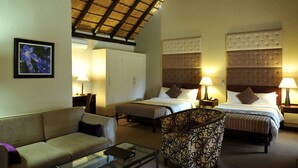 Quarto de hotel com duas camas, um sofá, uma poltrona, uma mesinha e teto com telhado de palha.