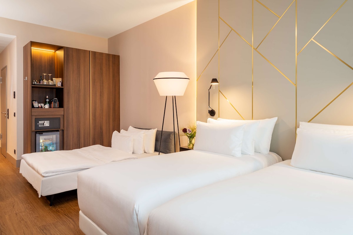 Quarto de hotel com duas camas, um guarda-roupa, uma mesinha de cabeceira com abajur e um frigorífico pequeno.