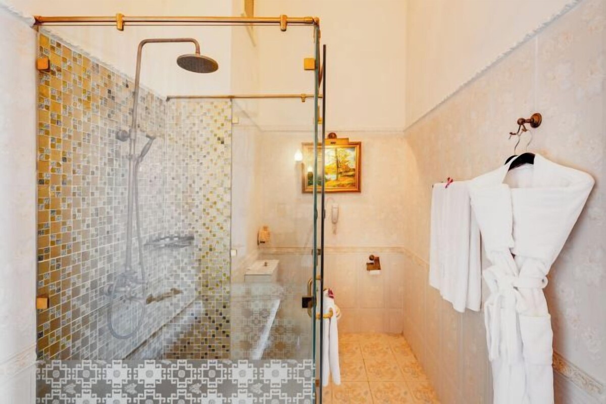 Um chuveiro com porta de vidro, um roupão branco pendurado em um gancho e uma parede revestida de azulejos com padrão em mosaico.