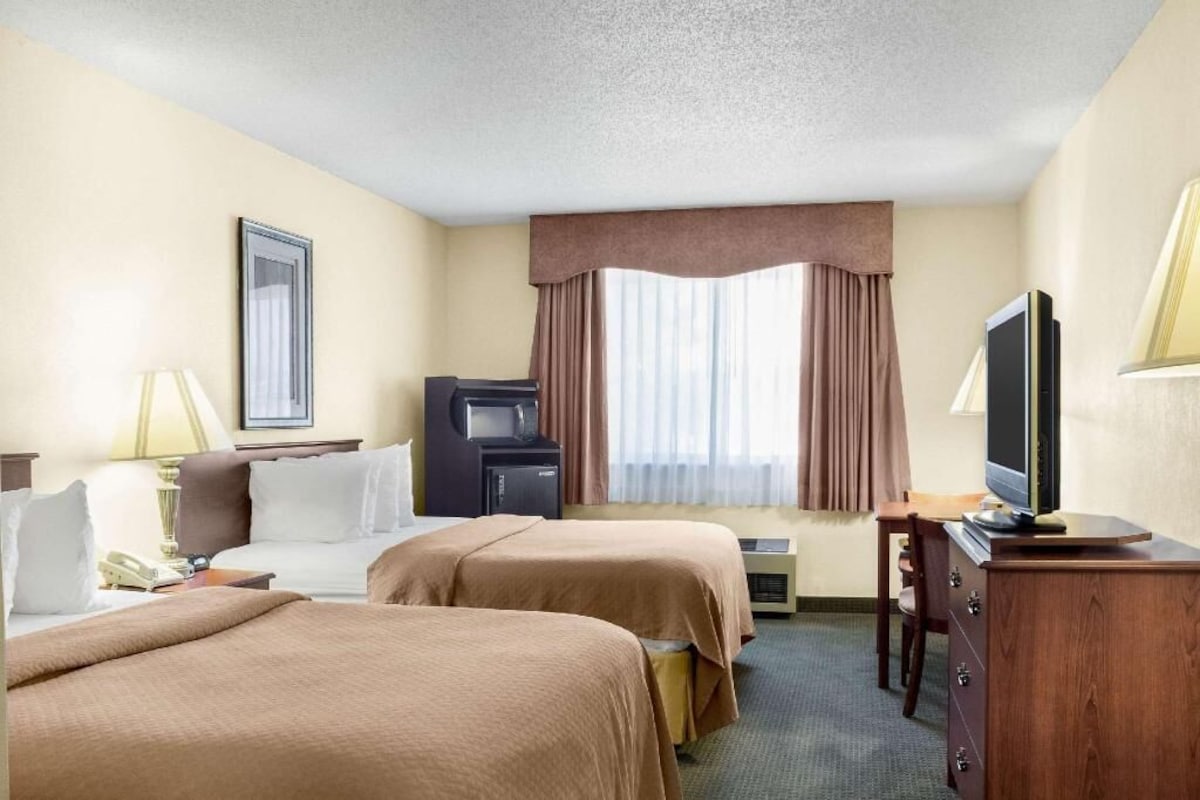 Quarto de hotel com duas camas, uma escrivaninha, televisão, telefone e janela com cortinas.