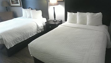 Quarto de hotel com duas camas, cada uma com roupa de cama branca e cabeceiras. Há uma mesa de cabeceira entre as camas com uma luminária e um relógio.