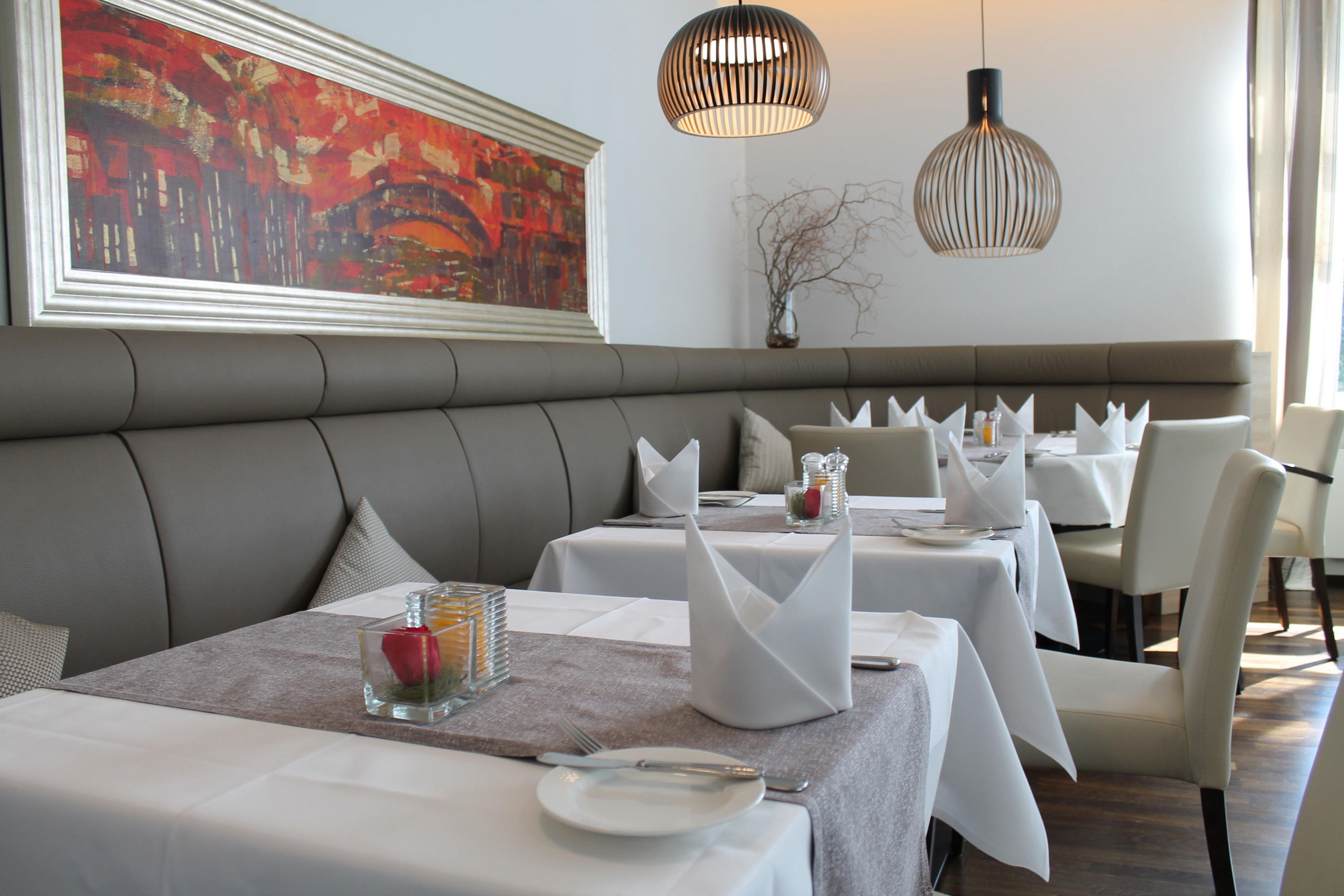 Interior de restaurante com área de bancos alongada, mesas com toalhas brancas e iluminação pendente moderna.