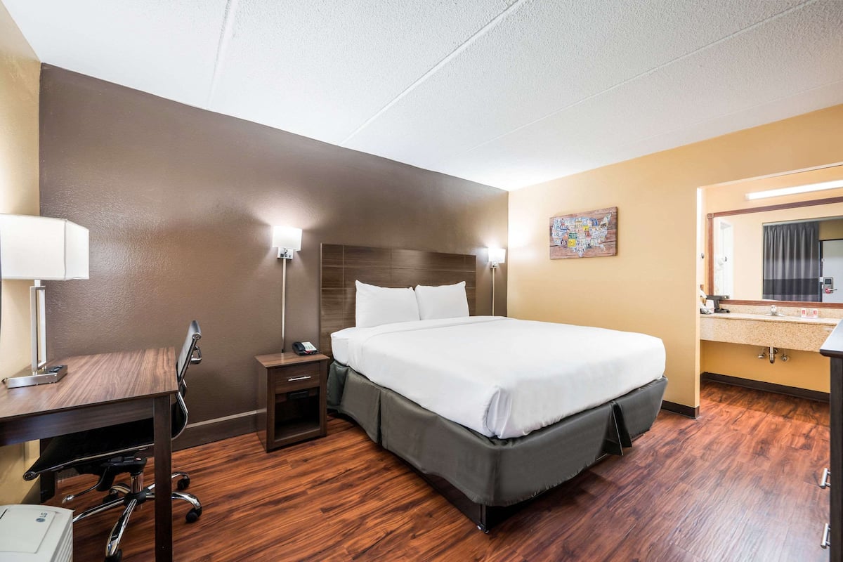 Quarto de hotel com uma cama grande, uma escrivaninha com luminária, mesa de cabeceira e banheiro com espelho e pia.