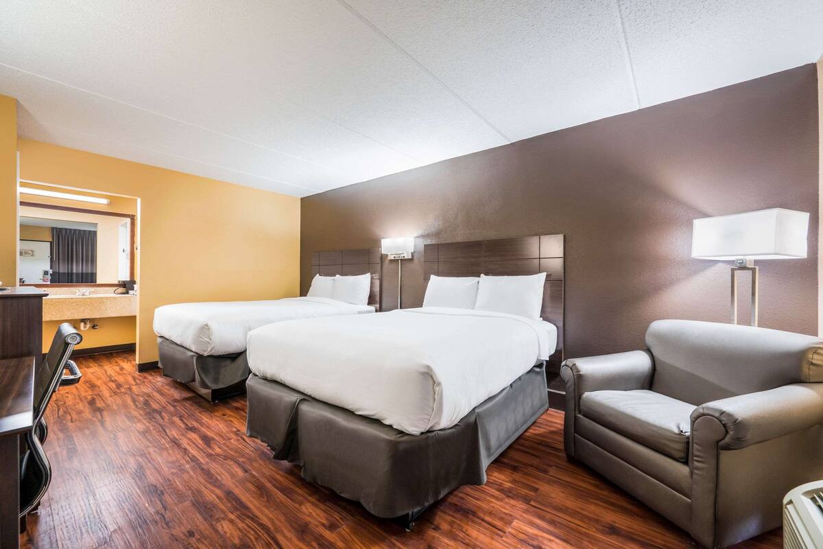 Quarto de hotel com duas camas, uma escrivaninha, uma cadeira e uma luminária.