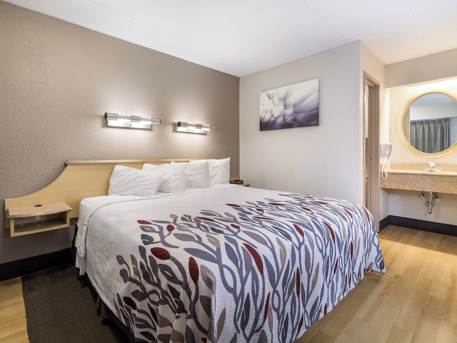 Quarto de hotel com cama, mesa de cabeceira, luminárias na parede, um quadro e banheiro com espelho e pia.