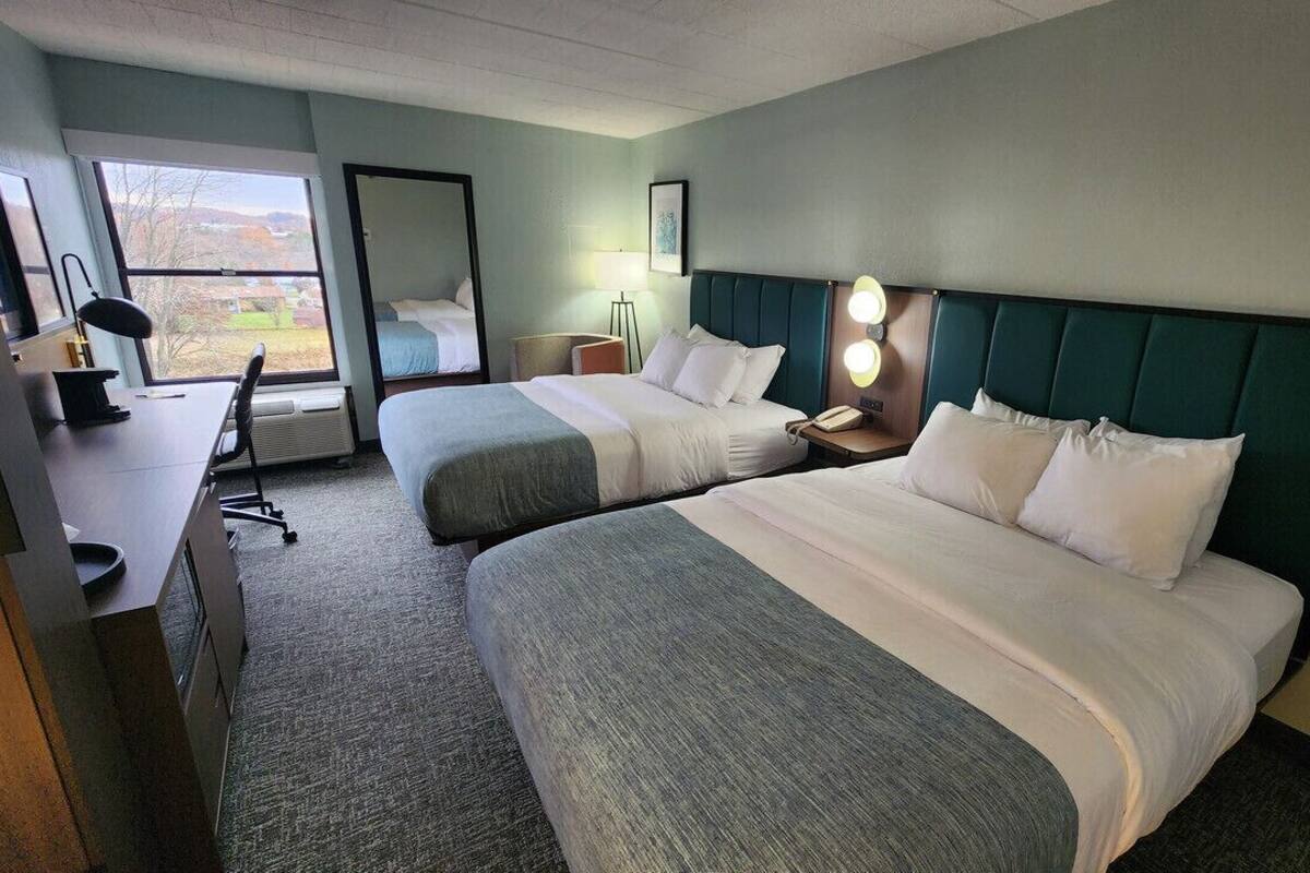 Quarto de hotel com duas camas, uma escrivaninha, uma cadeira, um abajur e janela com vista.