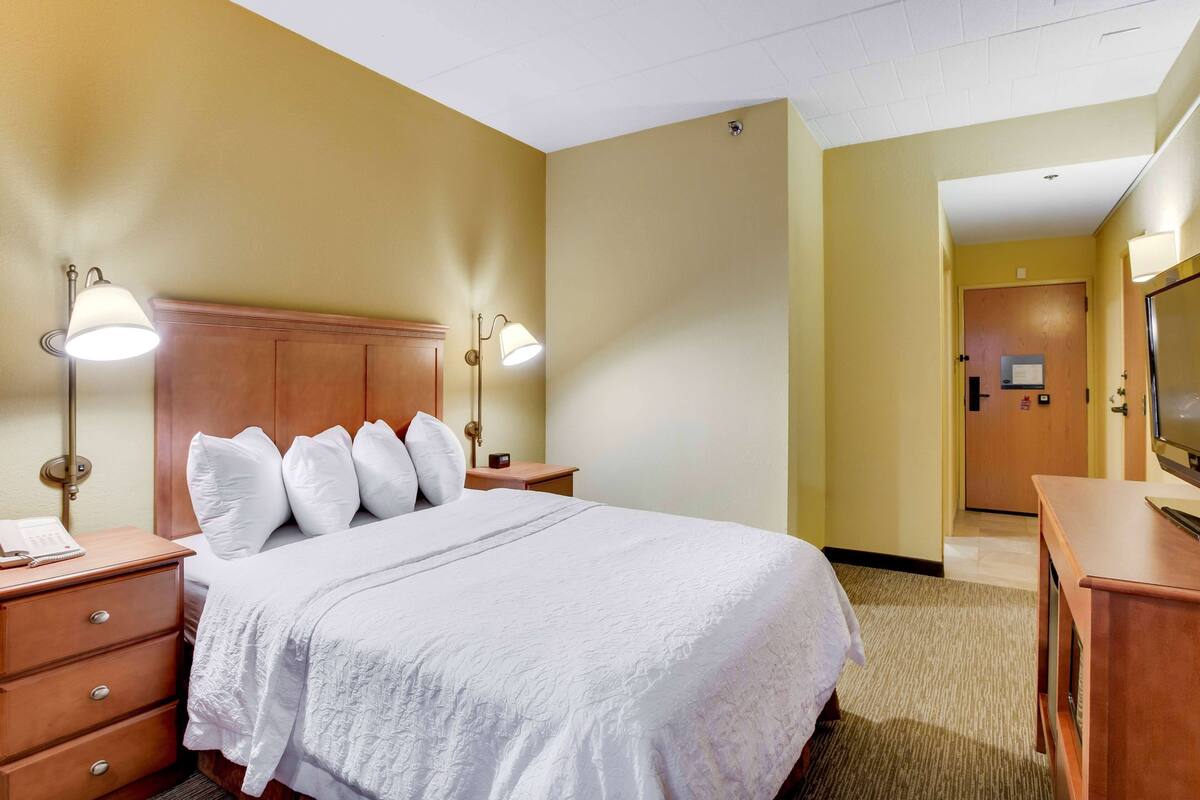 Quarto de hotel com cama, duas luminárias de cabeceira, mesa de cabeceira de madeira, escrivaninha com televisão e porta que dá para outro quarto.