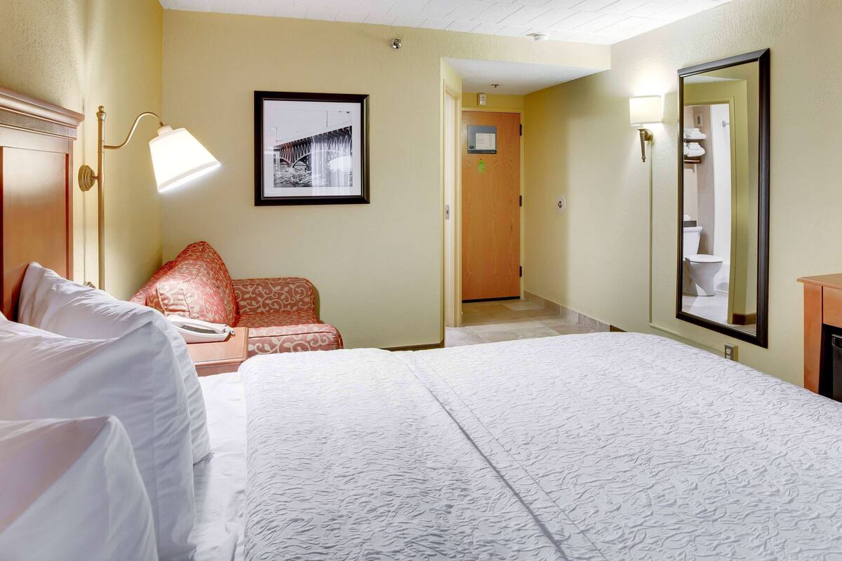 Quarto de hotel com uma cama grande, uma mesa de cabeceira com luminária, uma cadeira, um quadro na parede e uma porta que dá para um banheiro.