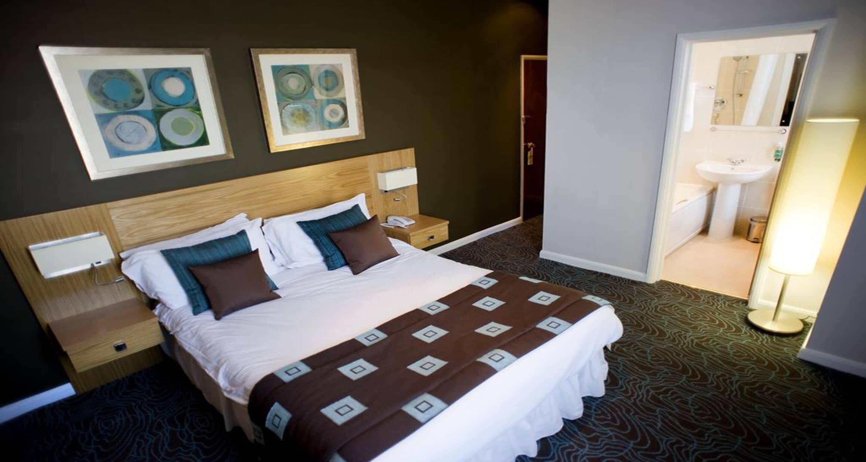 Quarto de hotel com uma cama grande, duas mesinhas de cabeceira, um criado-mudo, telefone fixado na parede e uma porta para o banheiro.