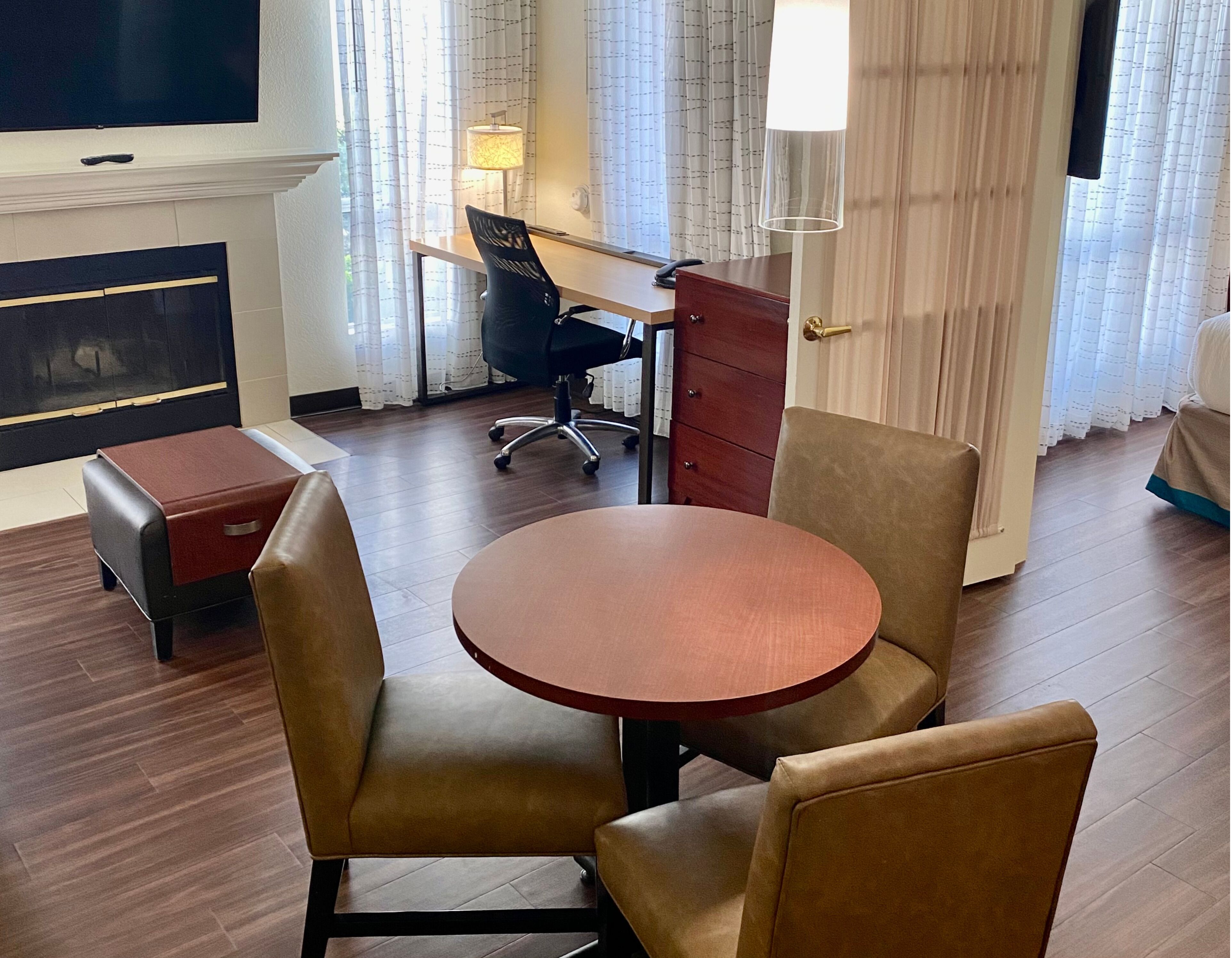 Quarto de hotel com uma mesa redonda de madeira, duas cadeiras bege, uma cadeira de escritório preta, uma escrivaninha com uma luminária, uma TV fixada na parede e uma lareira.