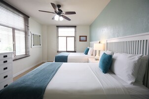 Quarto de hotel com duas camas, um ventilador de teto, janela com persianas, mesa de cabeceira com abajur e um quadro na parede.