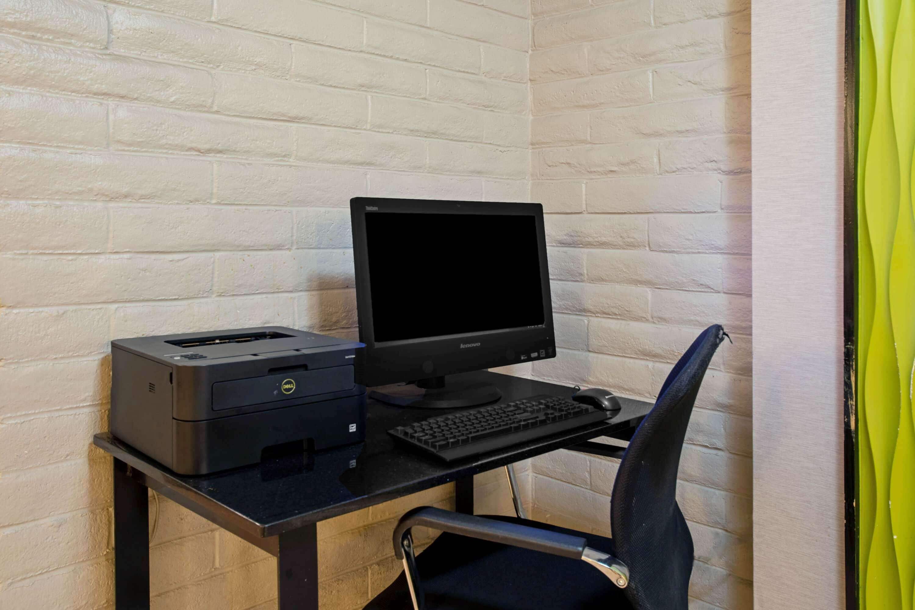 Uma mesa de escritório com monitor, teclado e impressora em frente a uma parede de tijolos.