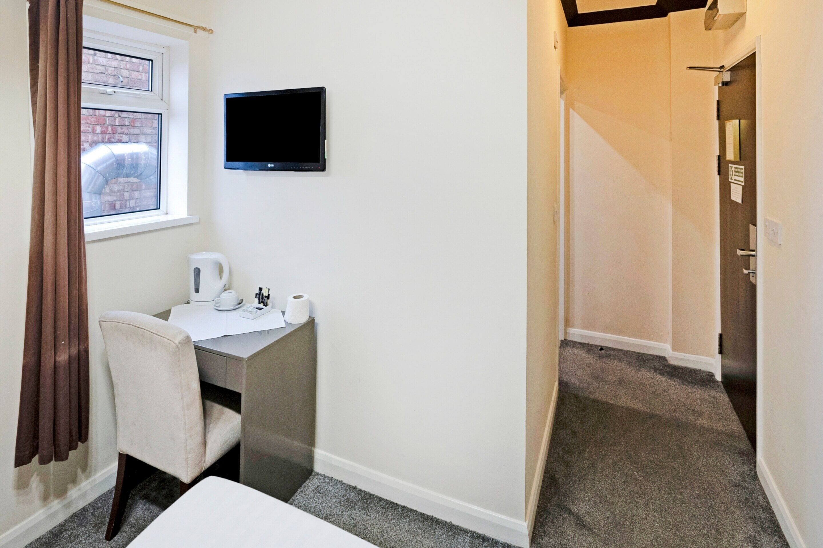 Quarto de hotel pequeno com escrivaninha, cadeira, televisão e janela com cortinas.