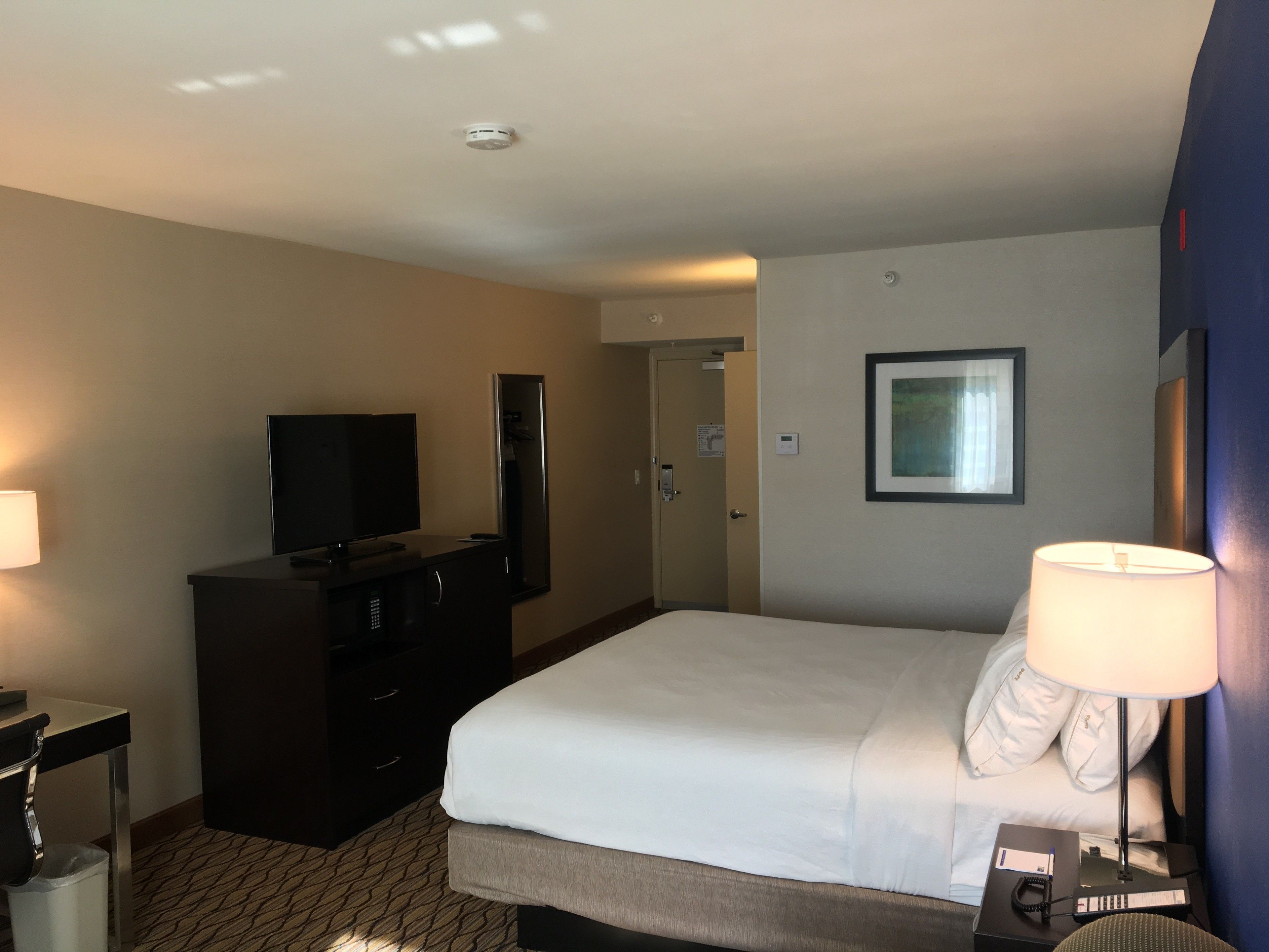 Quarto de hotel com cama, televisão em uma cômoda, abajur e porta que dá para outro espaço.