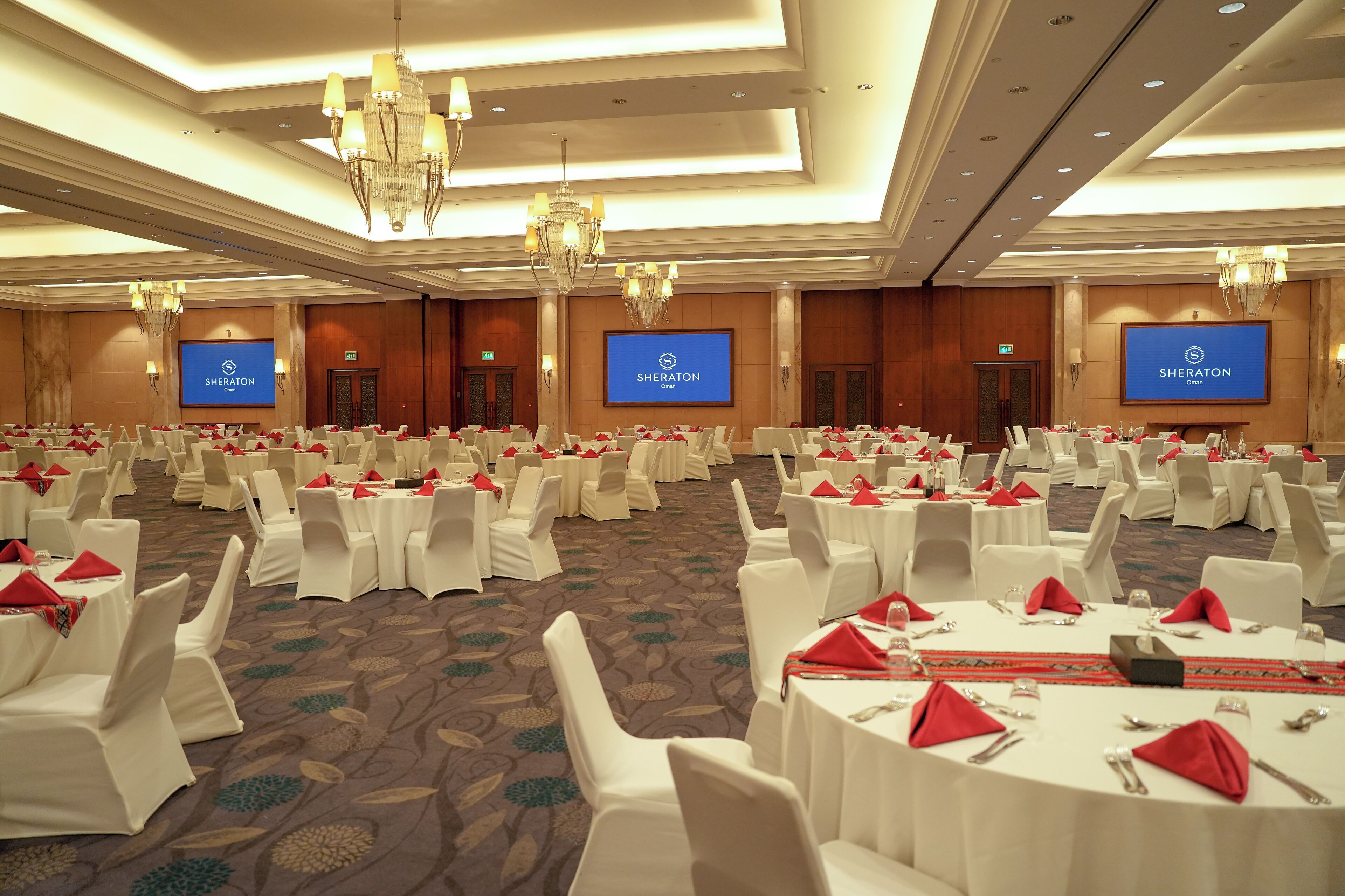 Salão de baile do Sheraton com diversas mesas redondas preparadas para refeições, cadeiras brancas, lustres e uma grande tela exibindo o logotipo do Sheraton.