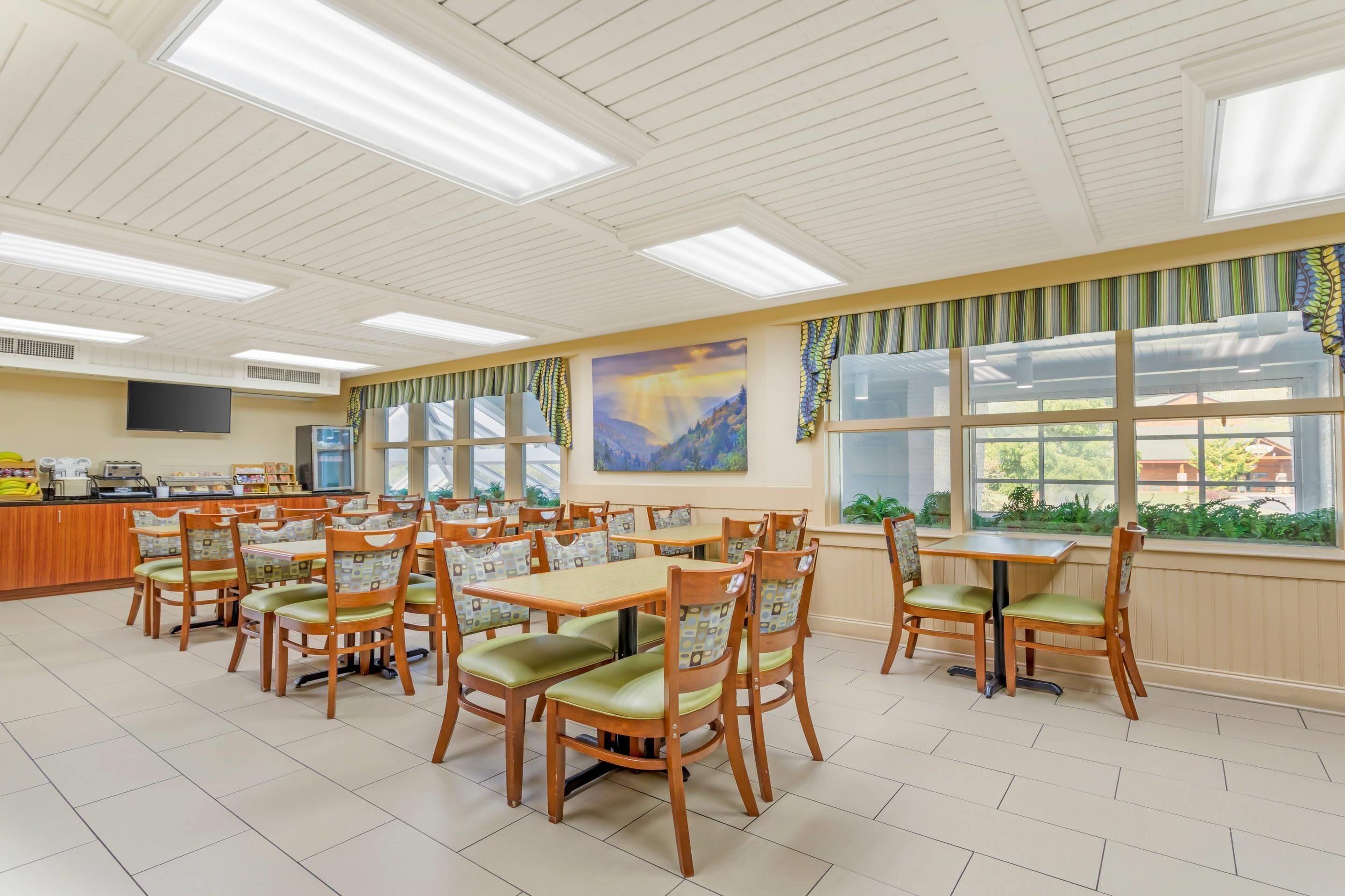 Uma área de refeições de hotel com várias mesas e cadeiras, um espaço de cozinha com eletrodomésticos e janelas amplas com cortinas.