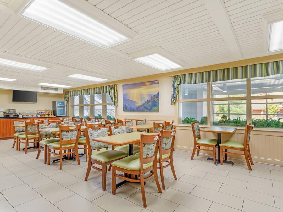 Uma área de refeições de hotel com várias mesas e cadeiras, um espaço de cozinha com eletrodomésticos e janelas amplas com cortinas.