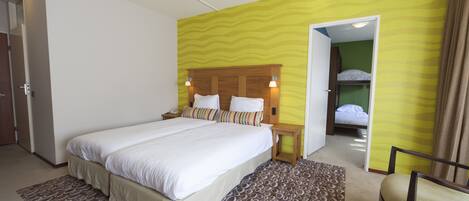 Quarto de hotel com cama grande, mesa de cabeceira, cadeira e uma cama beliche no quarto ao lado.