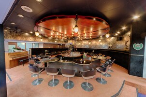 Uma área de bar com balcão circular, vários bancos e um piano ao fundo.