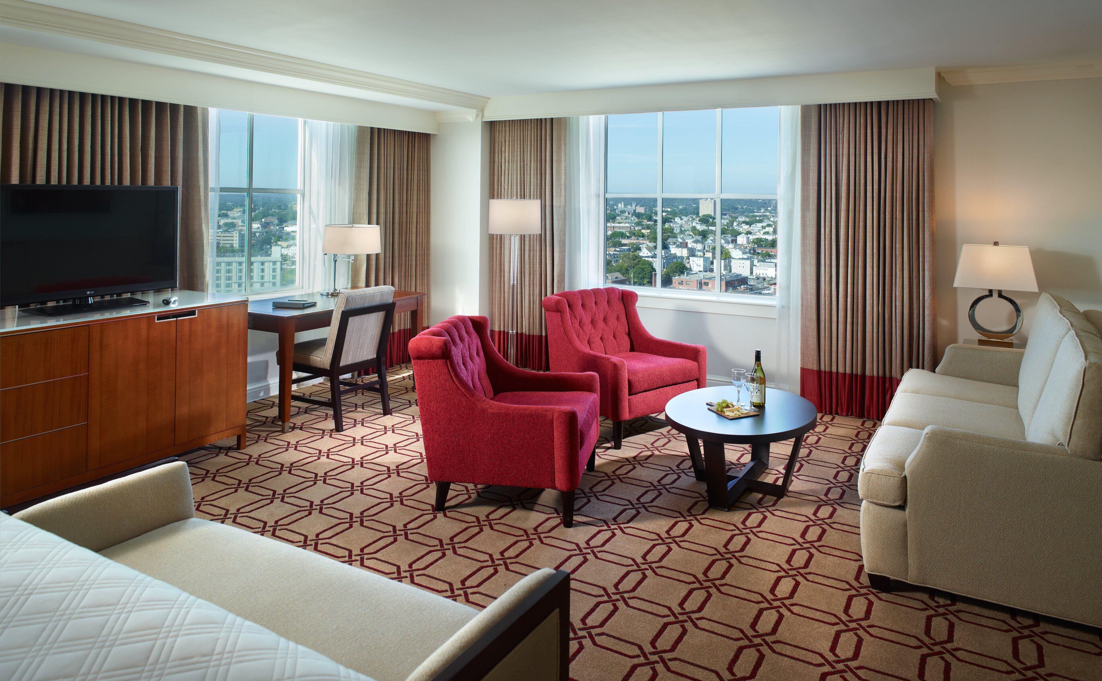 Quarto de hotel com uma cama, televisão, duas poltronas vermelhas, um sofá bege, uma mesinha com uma garrafa e copos, e vista para a cidade através de grandes janelas.