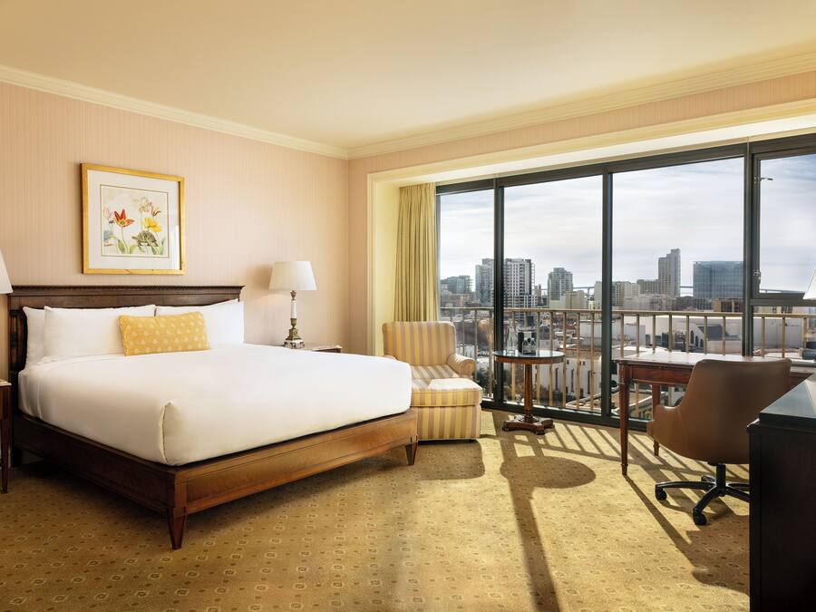Quarto de hotel com uma cama grande, uma escrivaninha, uma cadeira e vista para a cidade através de amplas janelas.