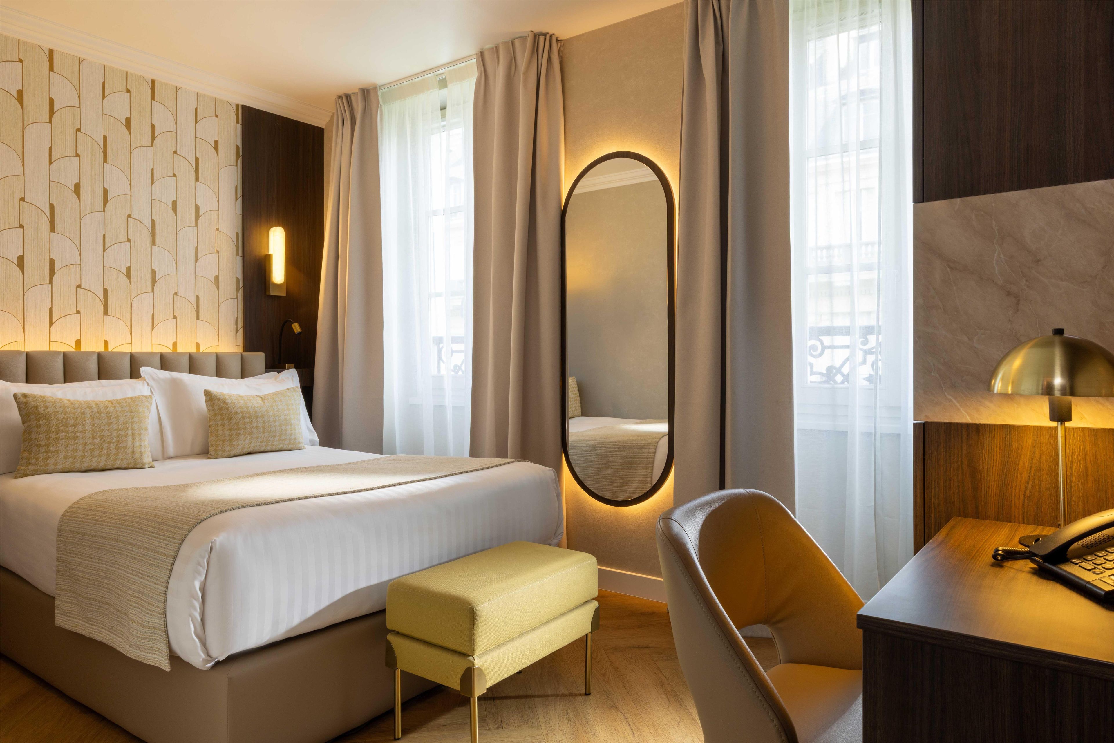 Quarto de hotel moderno com uma cama, escrivaninha, cadeira, espelho e parede com padrão geométrico.