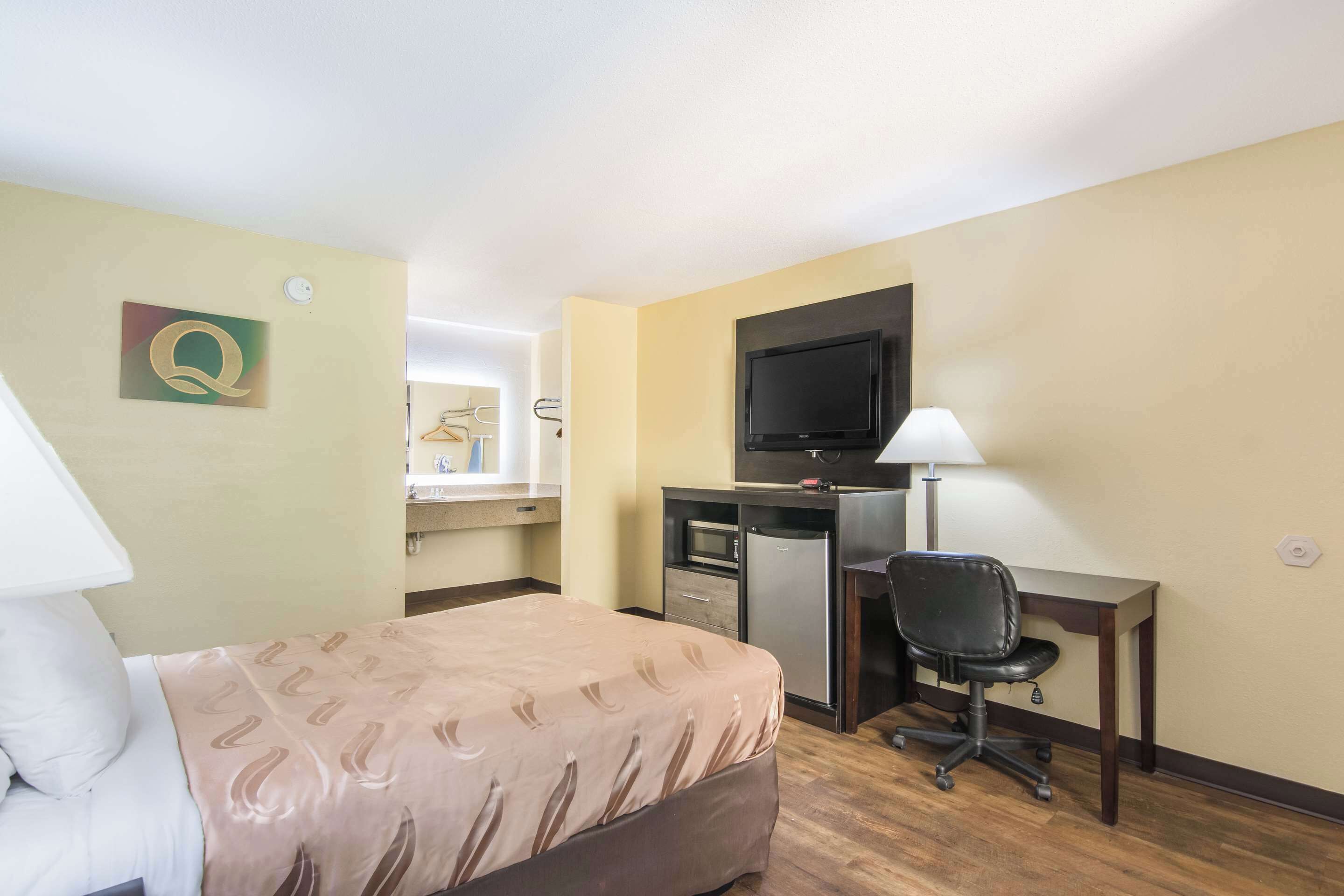 Quarto de hotel com cama, escrivaninha, cadeira, televisão e uma pequena cozinha equipada.
