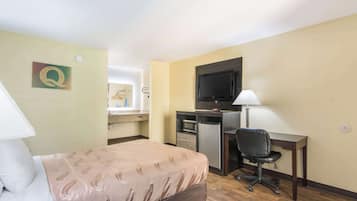 Quarto de hotel com cama, escrivaninha, cadeira, televisão e uma pequena cozinha equipada.
