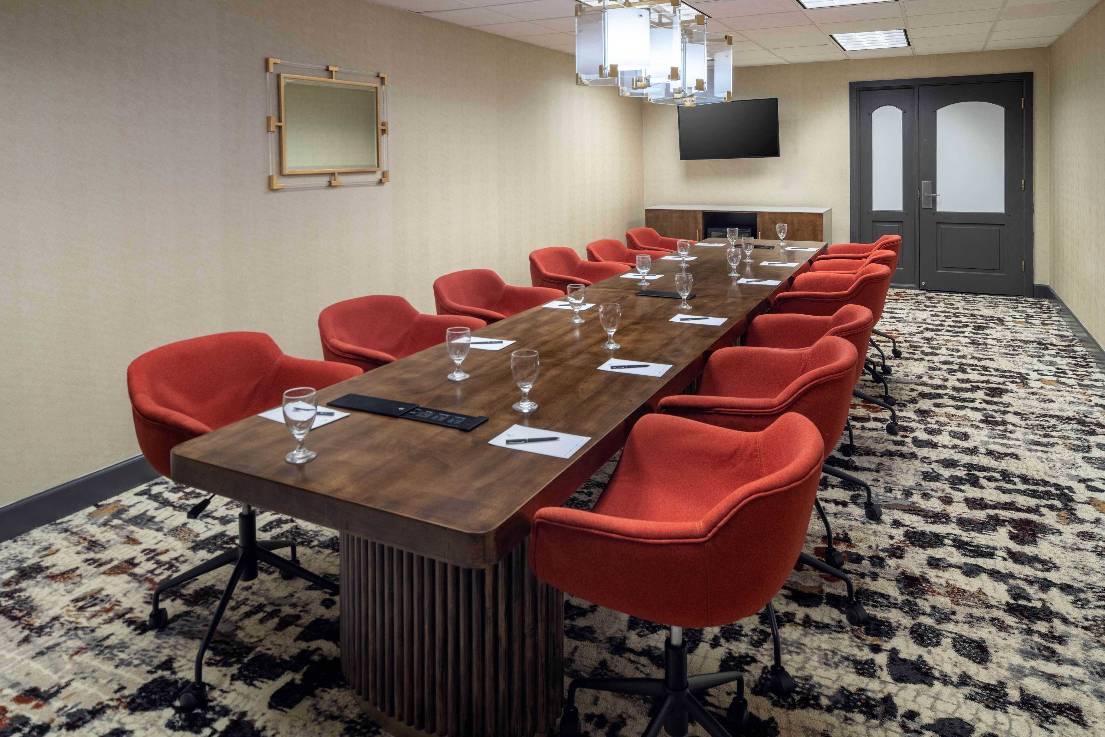 Uma sala de conferências com uma mesa comprida, cadeiras vermelhas, uma tela de projetor e uma televisão na parede.