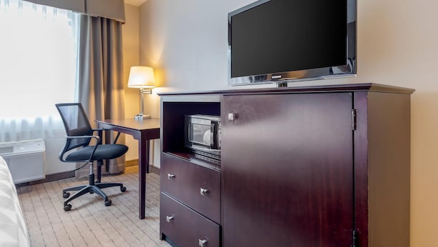 Quarto de hotel com televisão de tela plana presa a um móvel, uma mesa com luminária, uma cadeira e uma cama.