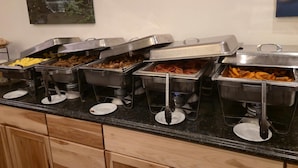 Buffet com diversos alimentos em recipientes metálicos, um armário de madeira abaixo e uma pintura de cachoeira na parede.