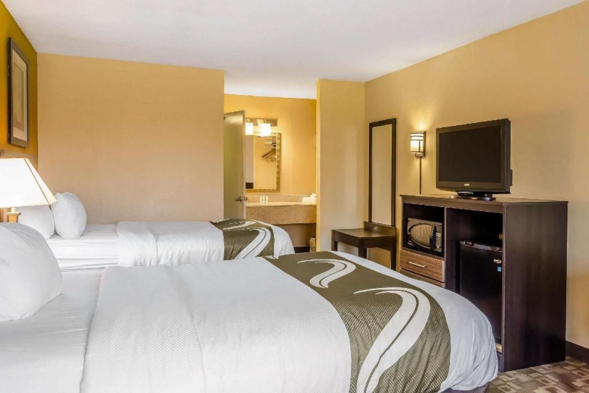 Quarto de hotel com duas camas, televisão, espelho e banheiro visível ao fundo.