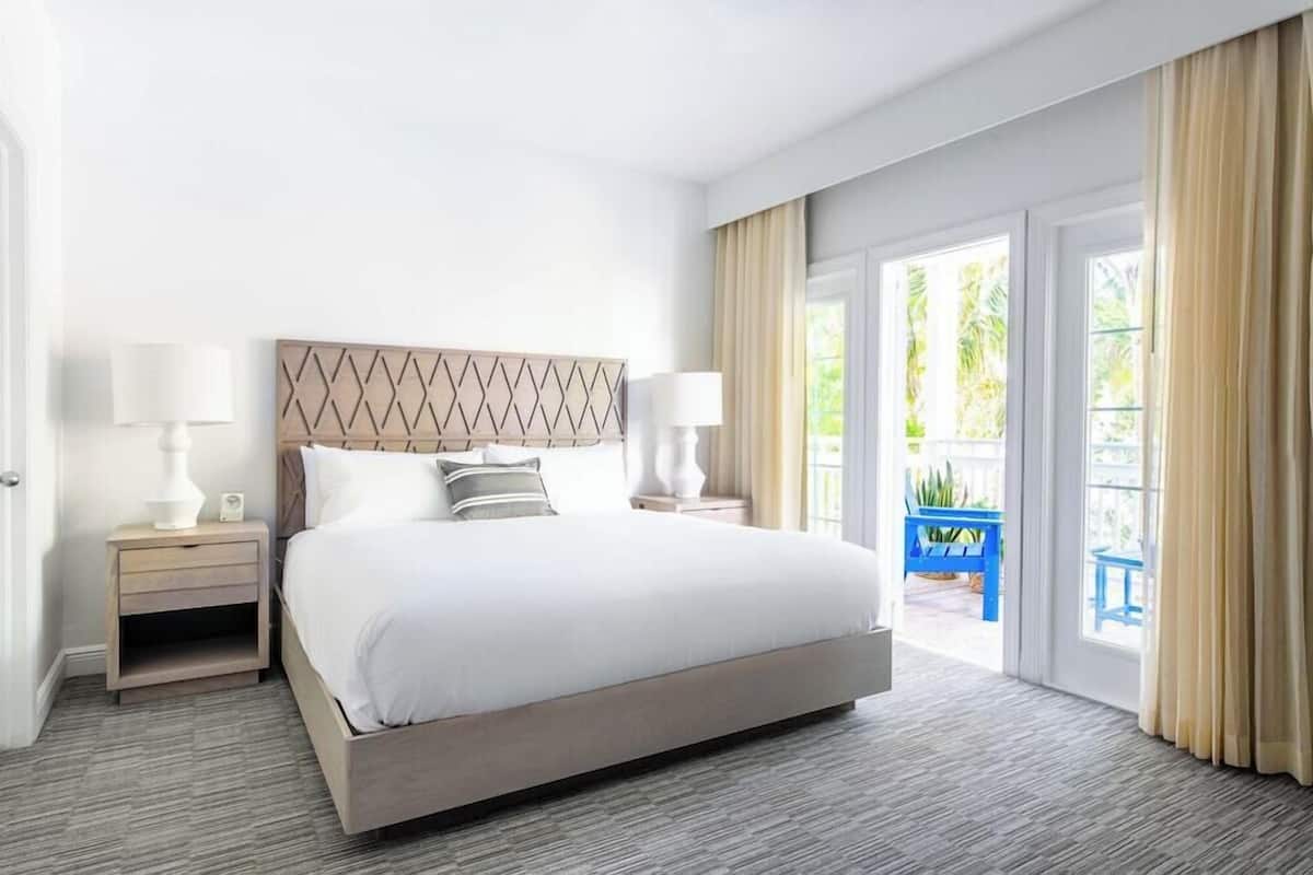 Quarto de hotel com uma cama grande, duas mesas de cabeceira com luminárias, uma cabeceira de madeira e vista para o exterior através de uma porta de vidro deslizante.