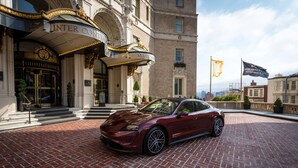 Um carro esportivo cor de vinho estacionado em frente à entrada imponente de um hotel com fachada de tijolos e detalhes ornamentados.