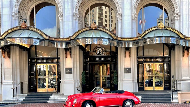 Um carro conversível vermelho clássico estacionado em frente a um hotel imponente com arquitetura ornamentada e várias entradas.