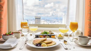 Mesa de café da manhã com pratos de comida, copos de suco de laranja e vista panorâmica da cidade pela janela.