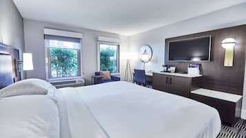 Quarto de hotel com uma cama grande, uma escrivaninha com computador, televisão de tela plana e janela com vista para vegetação.