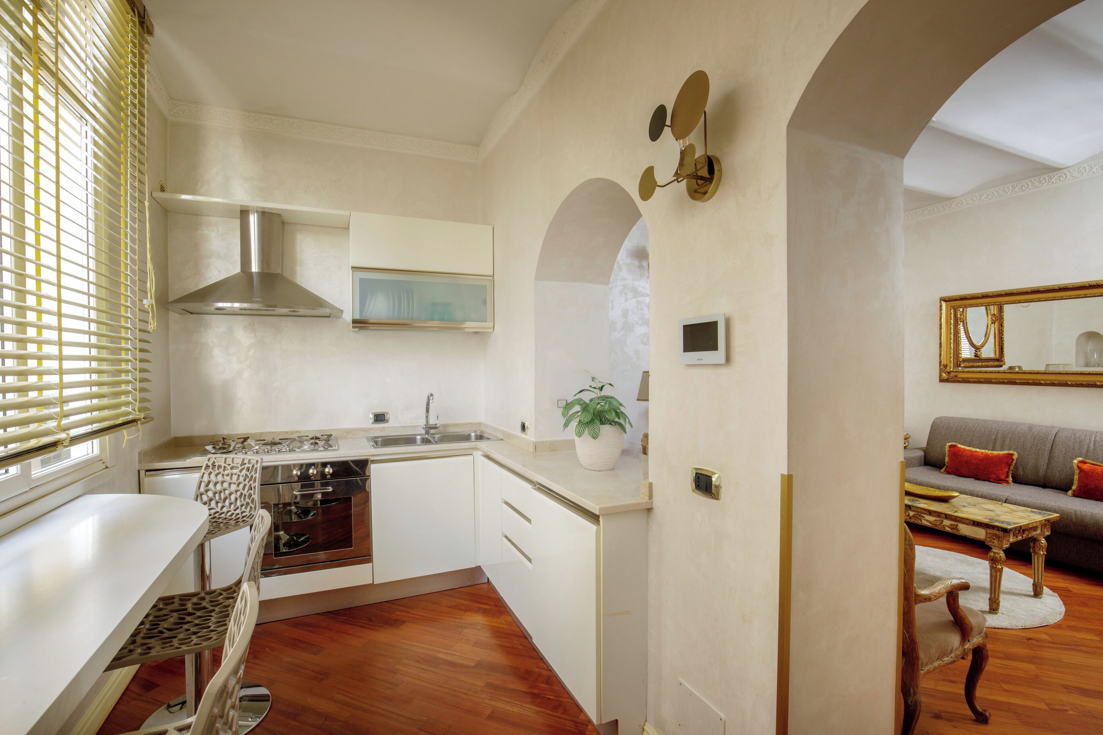 Cozinha moderna com armários brancos, coifa de aço inoxidável e forno embutido. Há uma área de jantar com mesa e cadeiras brancas, e uma sala de estar com sofá e espelho.