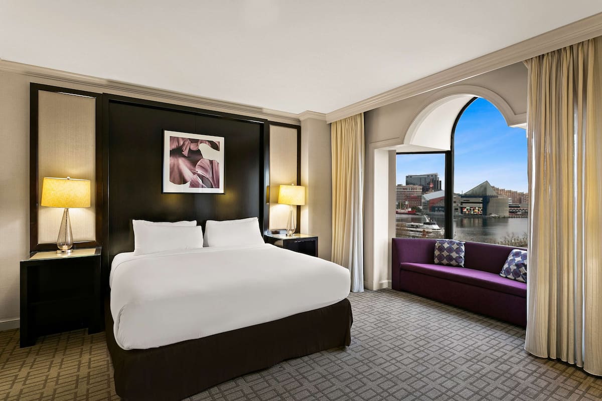 Quarto de hotel com uma cama grande, duas luminárias de cabeceira, um sofá roxo e vista para um horizonte urbano através de uma janela em arco.