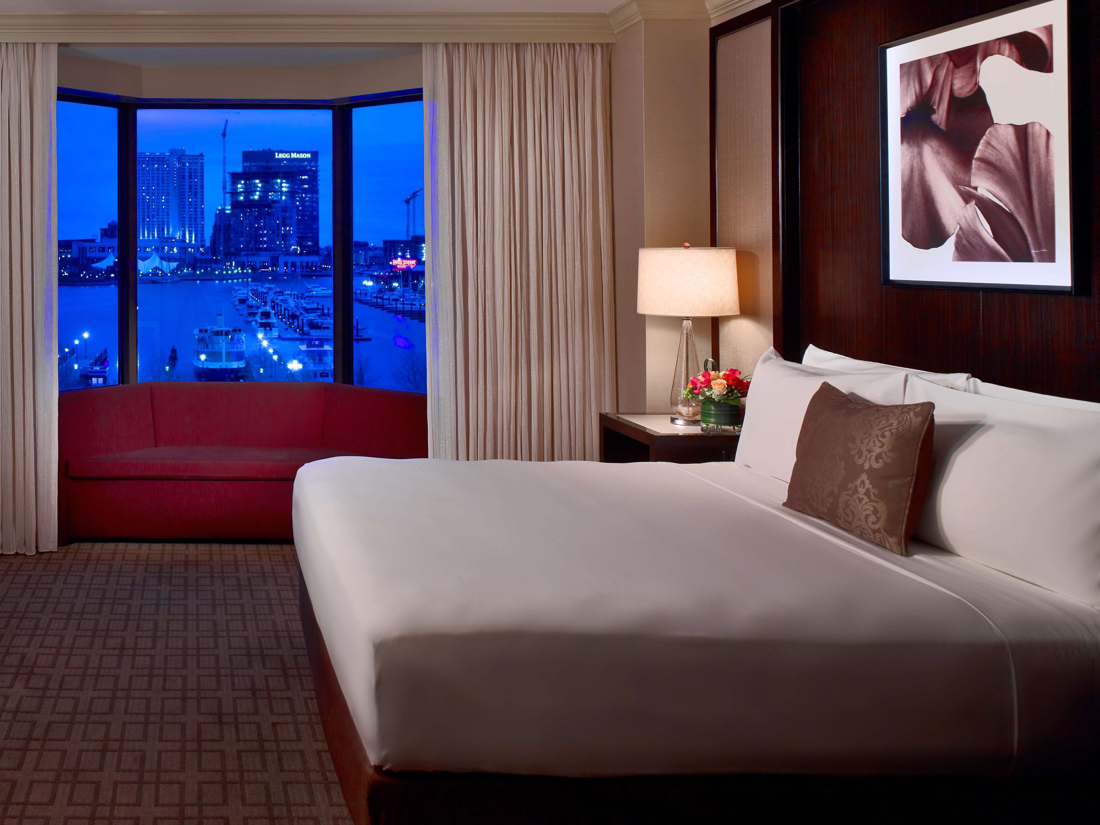 Quarto de hotel com uma cama grande, uma poltrona vermelha, uma mesinha de cabeceira com abajur e vista para a cidade ao entardecer.
