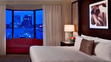 Quarto de hotel com uma cama grande, uma poltrona vermelha, uma mesinha de cabeceira com abajur e vista para a cidade ao entardecer.