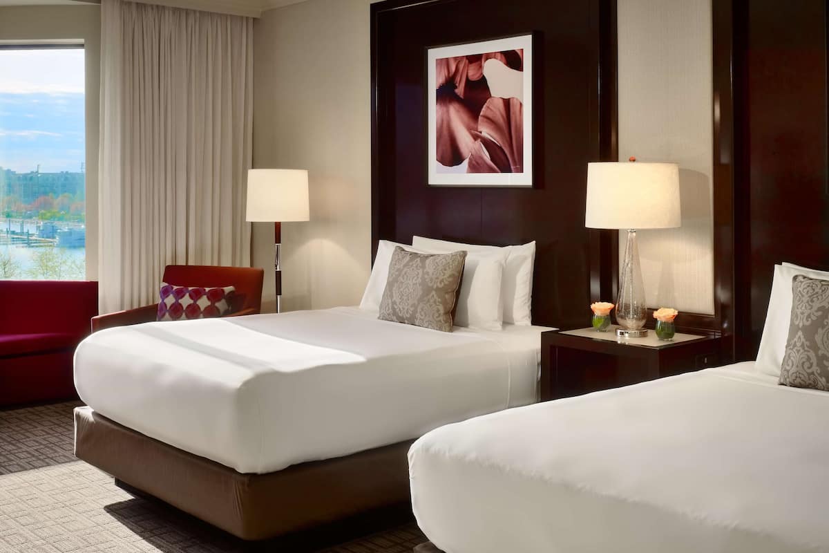 Quarto de hotel com duas camas, uma janela grande com vista para a cidade, uma cadeira vermelha e duas mesas de cabeceira com luminárias.