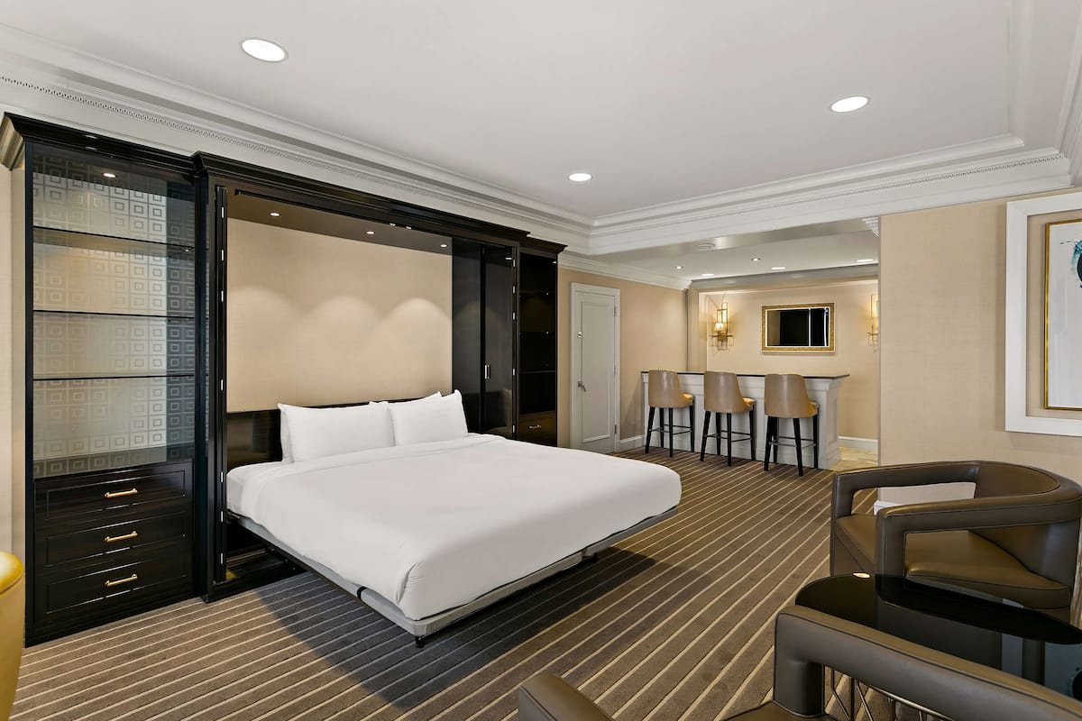 Quarto de hotel com uma cama grande, uma escrivaninha com duas cadeiras, um minibar e uma casa de banho com chuveiro e sanita.