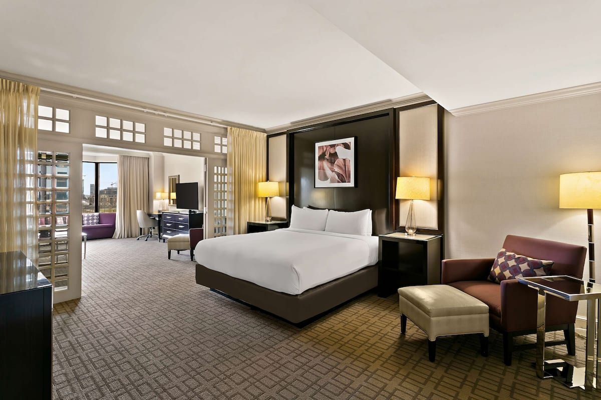 Quarto de hotel com uma cama grande, uma escrivaninha, uma cadeira e um sofá. Há duas luminárias nas mesas de cabeceira e um quadro na parede.