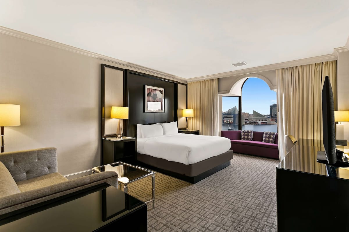 Quarto de hotel com uma cama grande, um sofá, uma escrivaninha e uma televisão. Há uma janela com vista para a paisagem urbana.