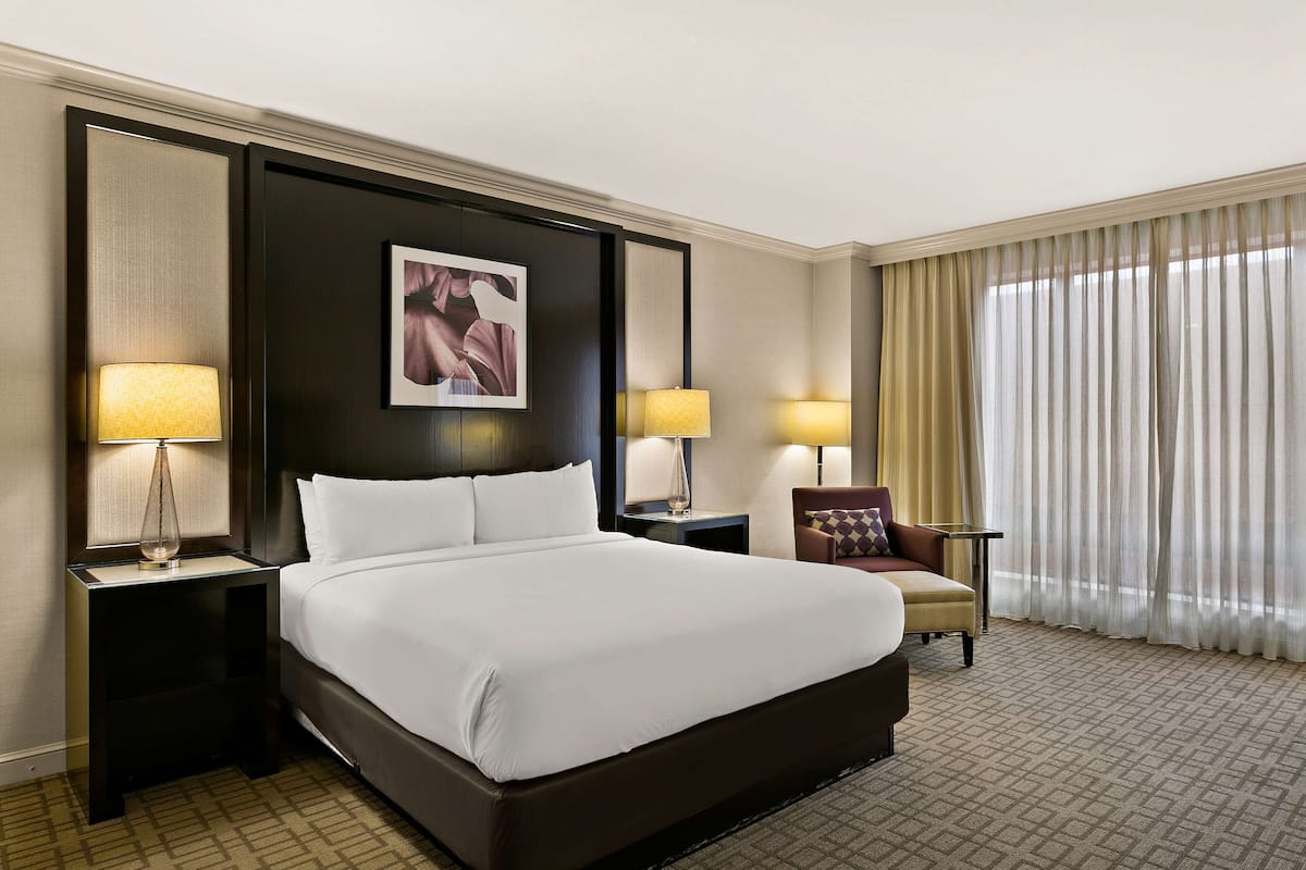 Quarto de hotel com uma cama grande, duas mesas de cabeceira com luminárias, uma cadeira e uma janela ampla com cortinas.