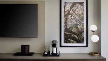 Quarto de hotel moderno com televisão de tela plana, fotografia emoldurada de uma árvore, cafeteira e abajur de dois braços.